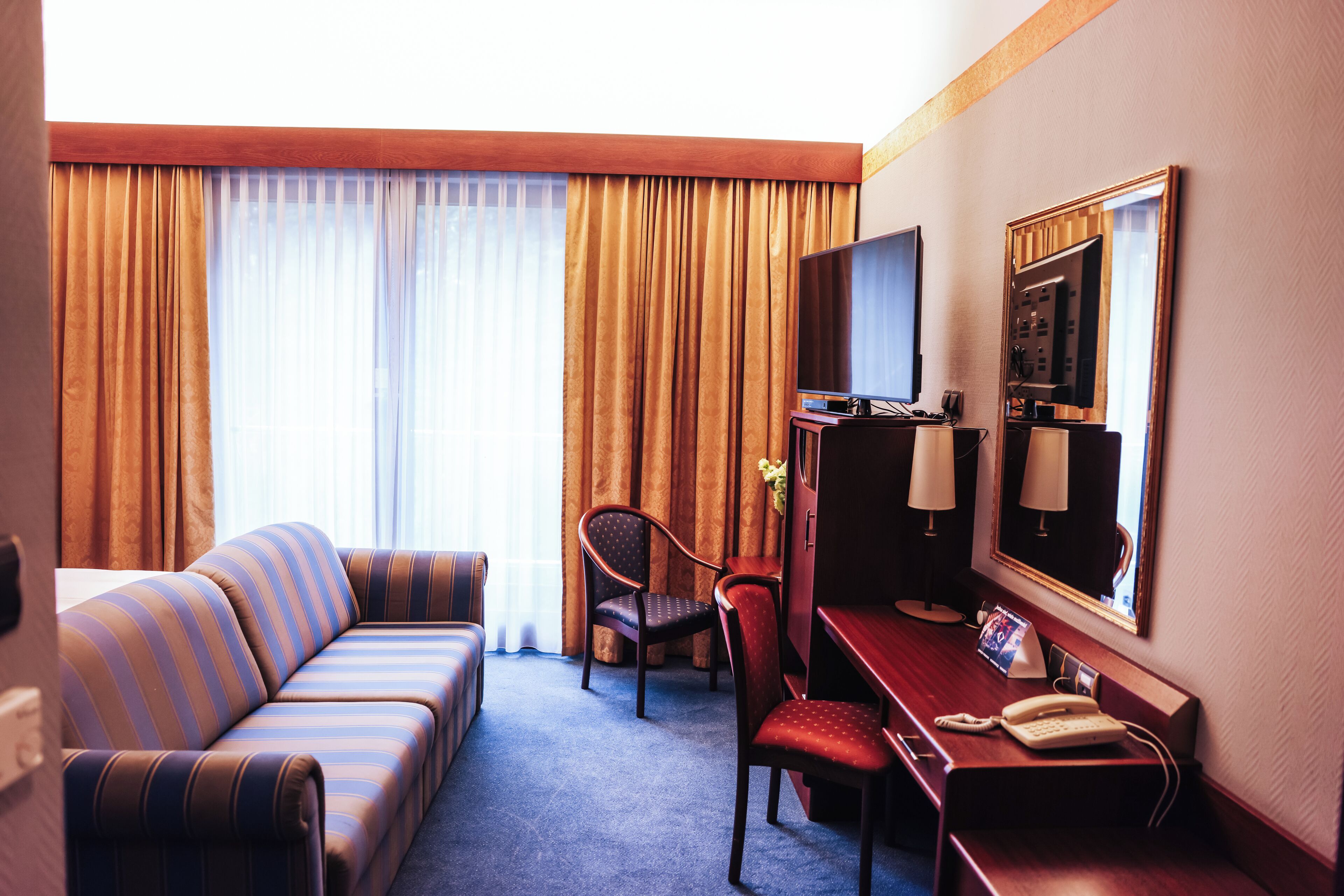 Quarto de hotel com sofá, escrivaninha com telefone, cadeira, televisão e uma grande janela com cortinas.