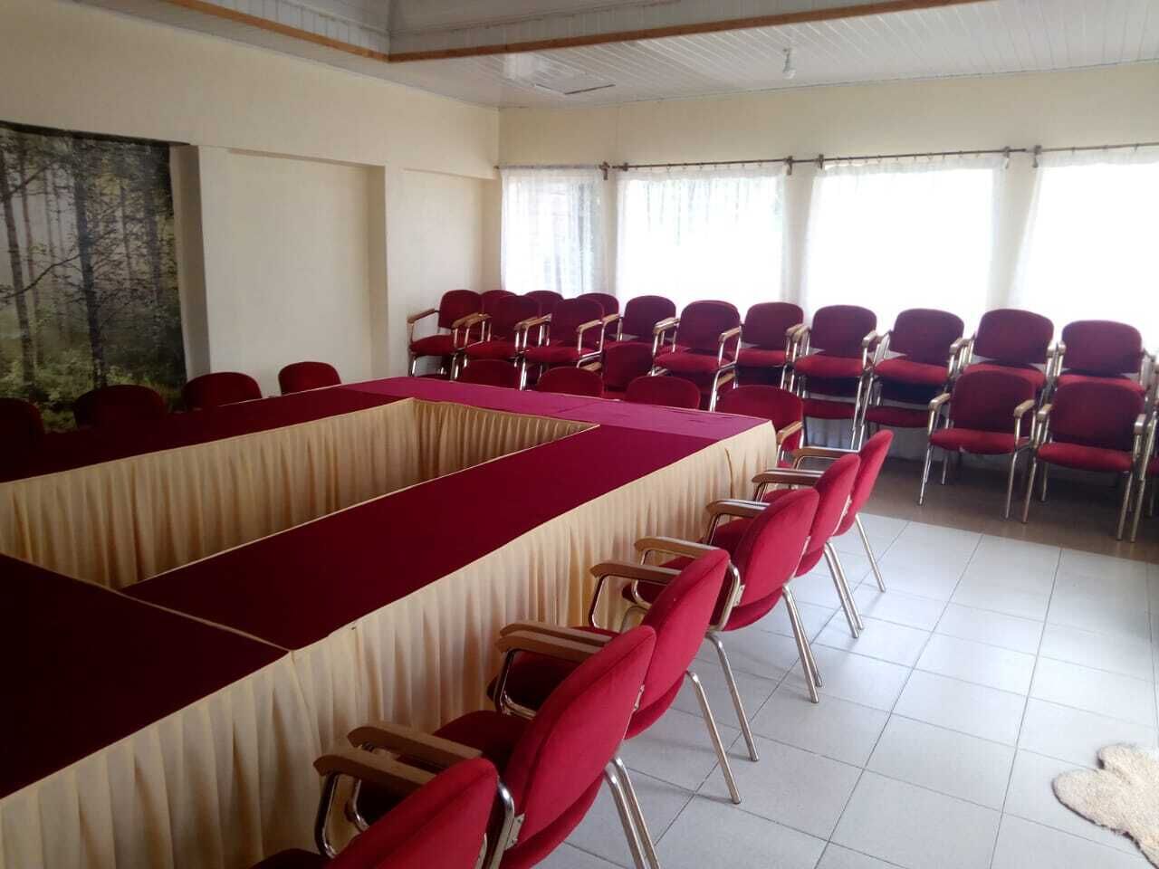 Uma sala de conferências com uma mesa comprida e cadeiras vermelhas, grandes janelas e um quadro na parede.