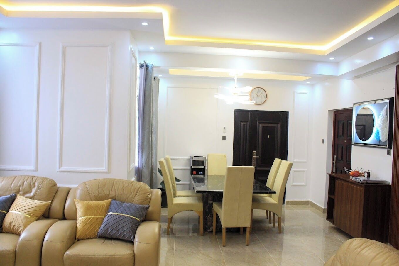 Uma sala de estar moderna com área de jantar, apresentando um conjunto de sofá, uma mesa de jantar com cadeiras e uma TV fixada na parede.