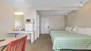 Quarto de hotel com cozinha compacta, uma cama de solteiro com colcha estampada, escrivaninha com cadeira e um espelho.