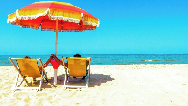 Duas cadeiras de praia sob um guarda-sol colorido numa praia de areia com um céu azul límpido.