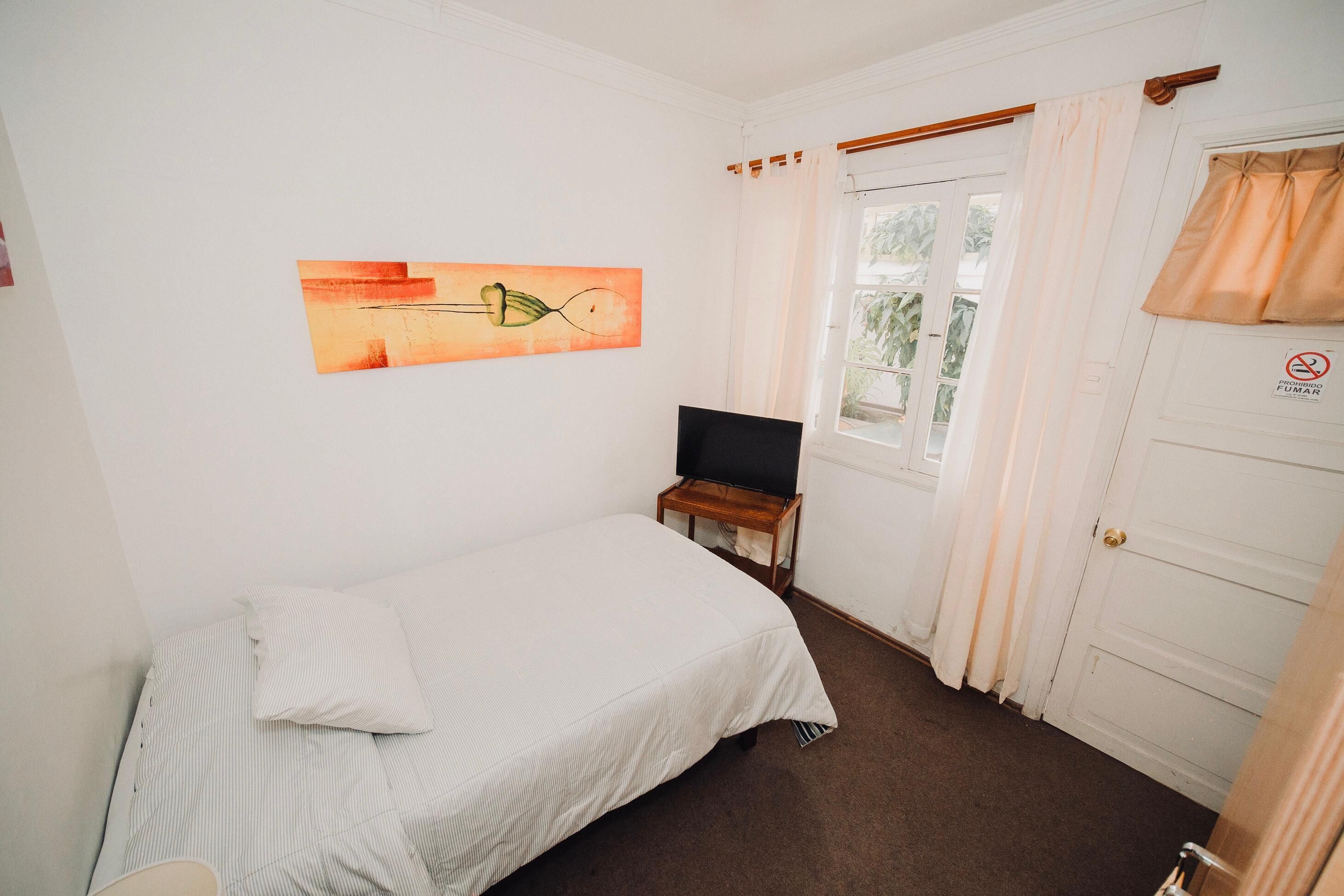 Quarto pequeno e bem organizado, com uma cama de solteiro, uma televisão sobre um suporte de madeira e uma janela com cortinas.