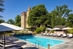 Uma piscina com espreguiçadeiras e guarda-sóis, uma torre de pedra e vegetação exuberante.