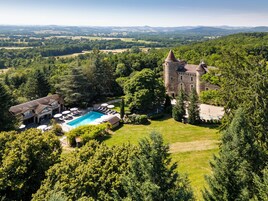 Um castelo com piscina, rodeado por vegetação exuberante e vista para colinas distantes.