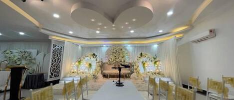 Salão de banquetes com uma longa mesa, cadeiras e palco decorado com flores.
