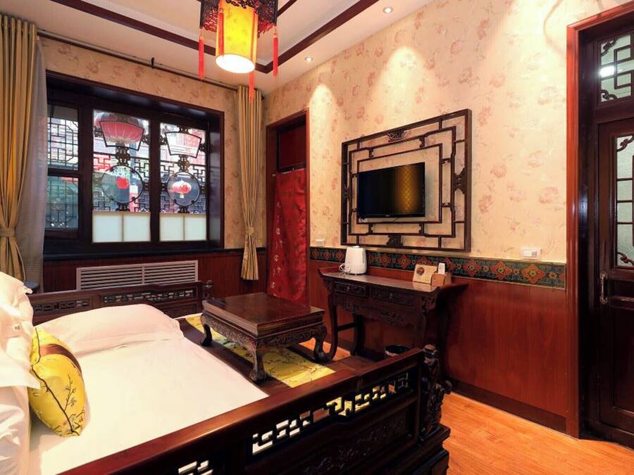 Quarto tradicional com uma cama, televisão na parede e janela com detalhes decorativos.