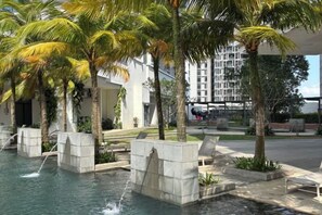 Uma área de piscina com palmeiras, espreguiçadeiras e um edifício moderno ao fundo.