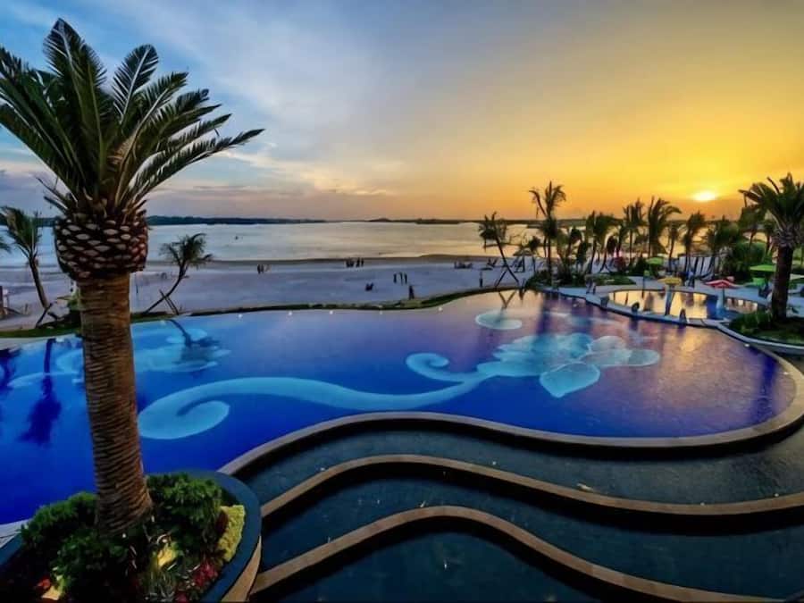 Uma piscina com design ondulado, palmeiras e vista para a praia ao pôr do sol.