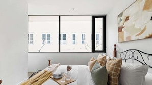 Quarto com uma janela grande, uma cama com roupa de cama branca e vista para edifícios.