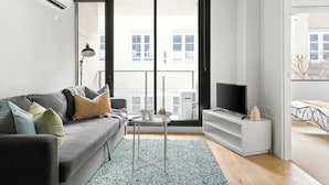 Uma sala de estar moderna com um sofá cinza, uma mesa redonda pequena, uma TV sobre um suporte branco e uma janela grande com vista para edifícios.