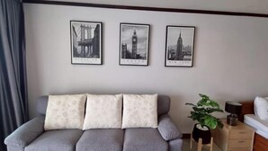 Um sofá cinzento com três almofadas estampadas, uma pequena mesa de cabeceira de madeira com uma planta, e três quadros em preto e branco com paisagens urbanas na parede.