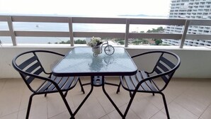 Uma varanda com mesa de vidro e duas cadeiras, um vaso de plantas e vista para o mar e edifícios.