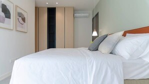 Uma cama bem arrumada com roupa de cama e almofadas brancas, um guarda-roupa com espelho e um quadro na parede.