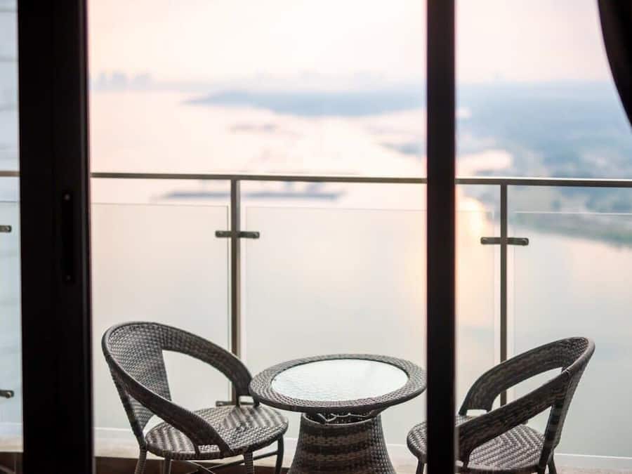 Varanda com cadeiras de vime e mesa de vidro, com vista para um corpo d'água calmo e o horizonte da cidade ao longe.