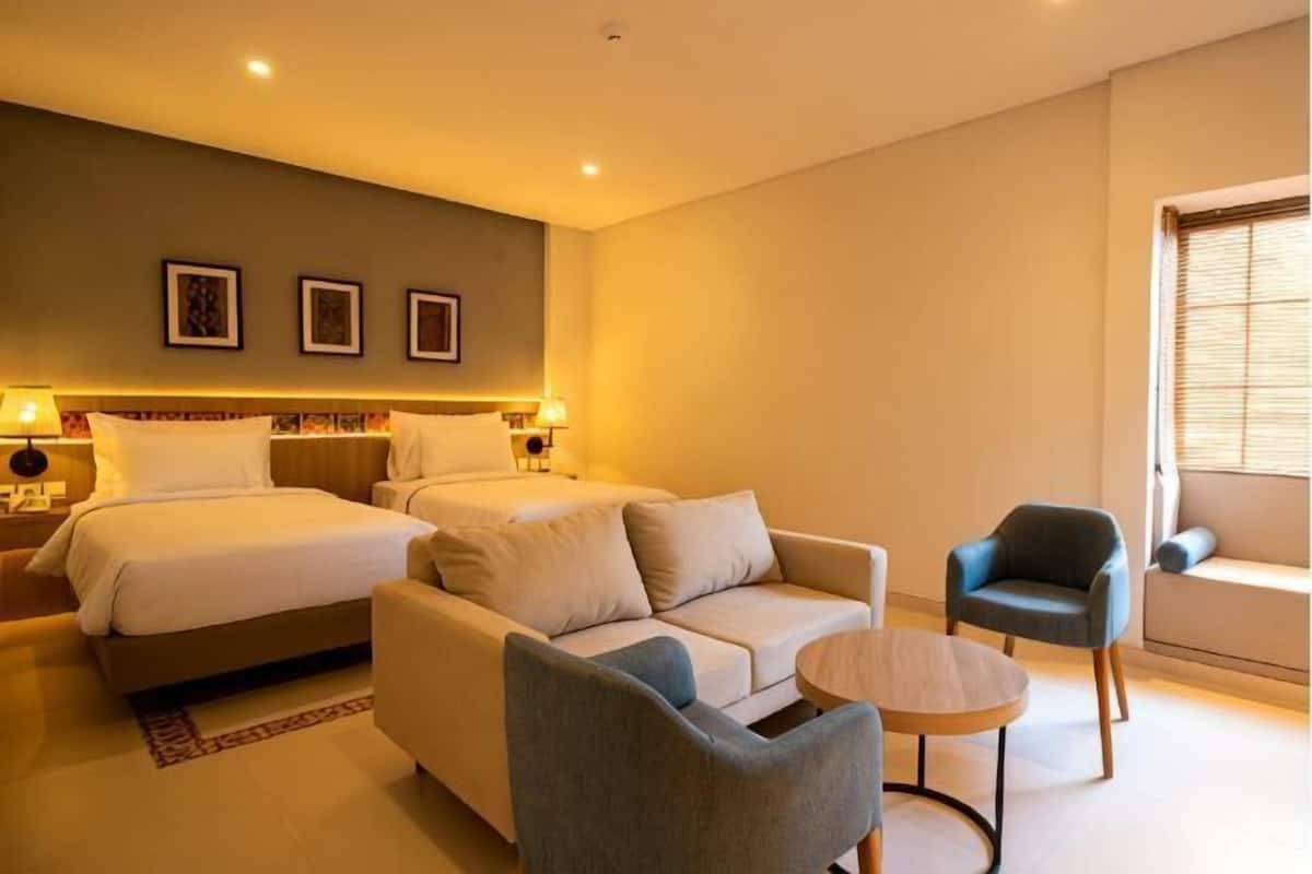 Quarto de hotel com uma cama, dois sofás, uma cadeira, uma mesa pequena e uma janela com persianas.