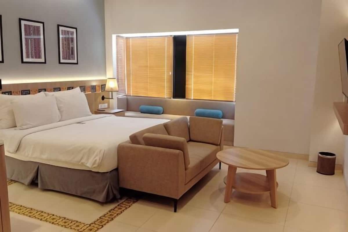 Quarto de hotel com cama, sofá, mesinha, janela com persianas e televisão fixada na parede.