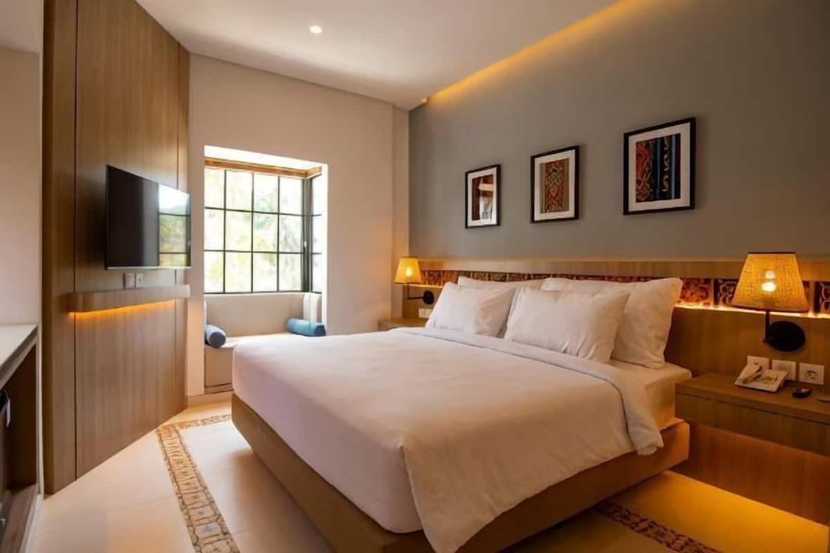 Quarto de hotel com uma cama grande, televisão na parede, mesa de cabeceira com abajur e janela que oferece iluminação natural.