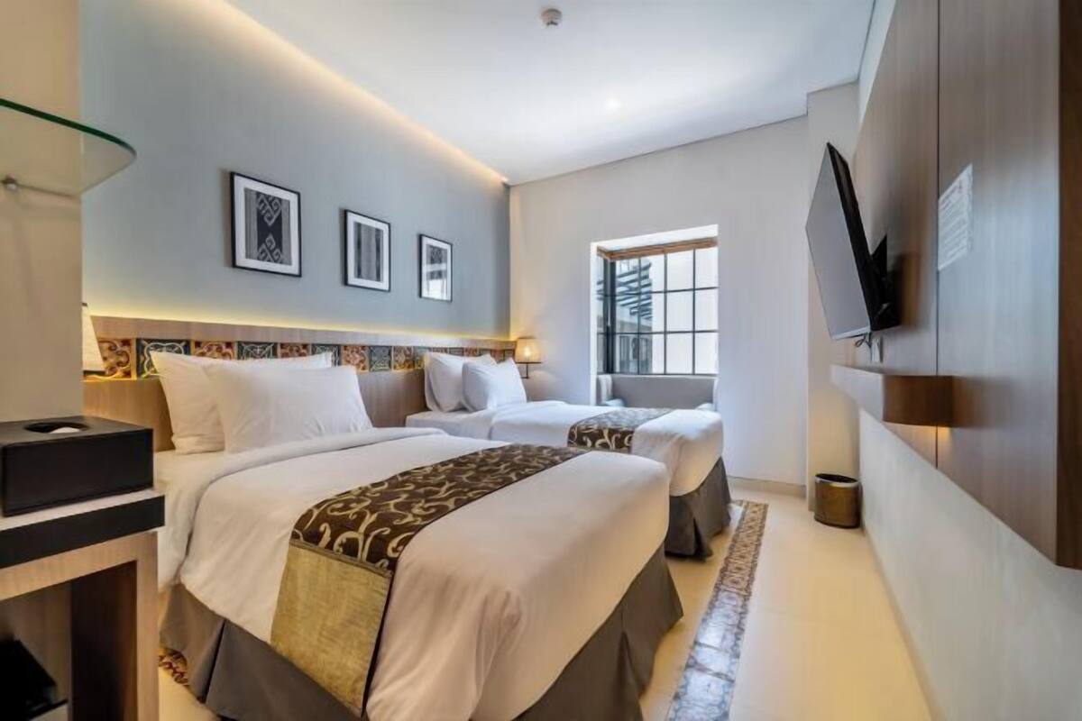 Quarto de hotel com duas camas, televisão na parede e janela com iluminação natural.