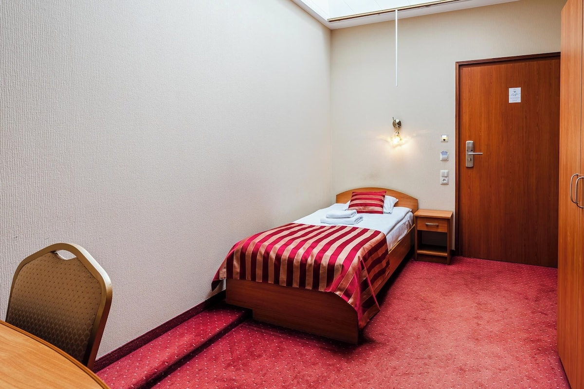 Quarto de hotel pequeno com uma cama de casal, uma mesinha de cabeceira de madeira, uma cadeira e uma porta.