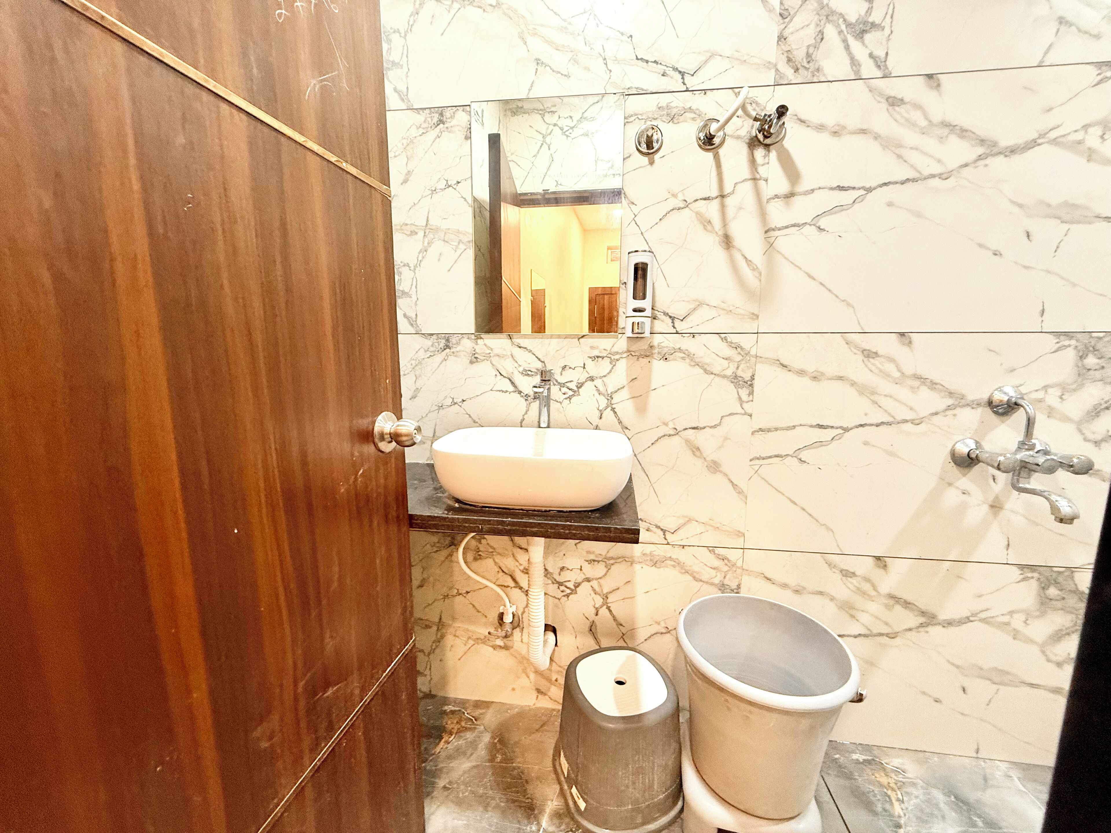 Casa de banho com porta de madeira, lavatório branco, parede com azulejos de mármore, espelho e sanita.