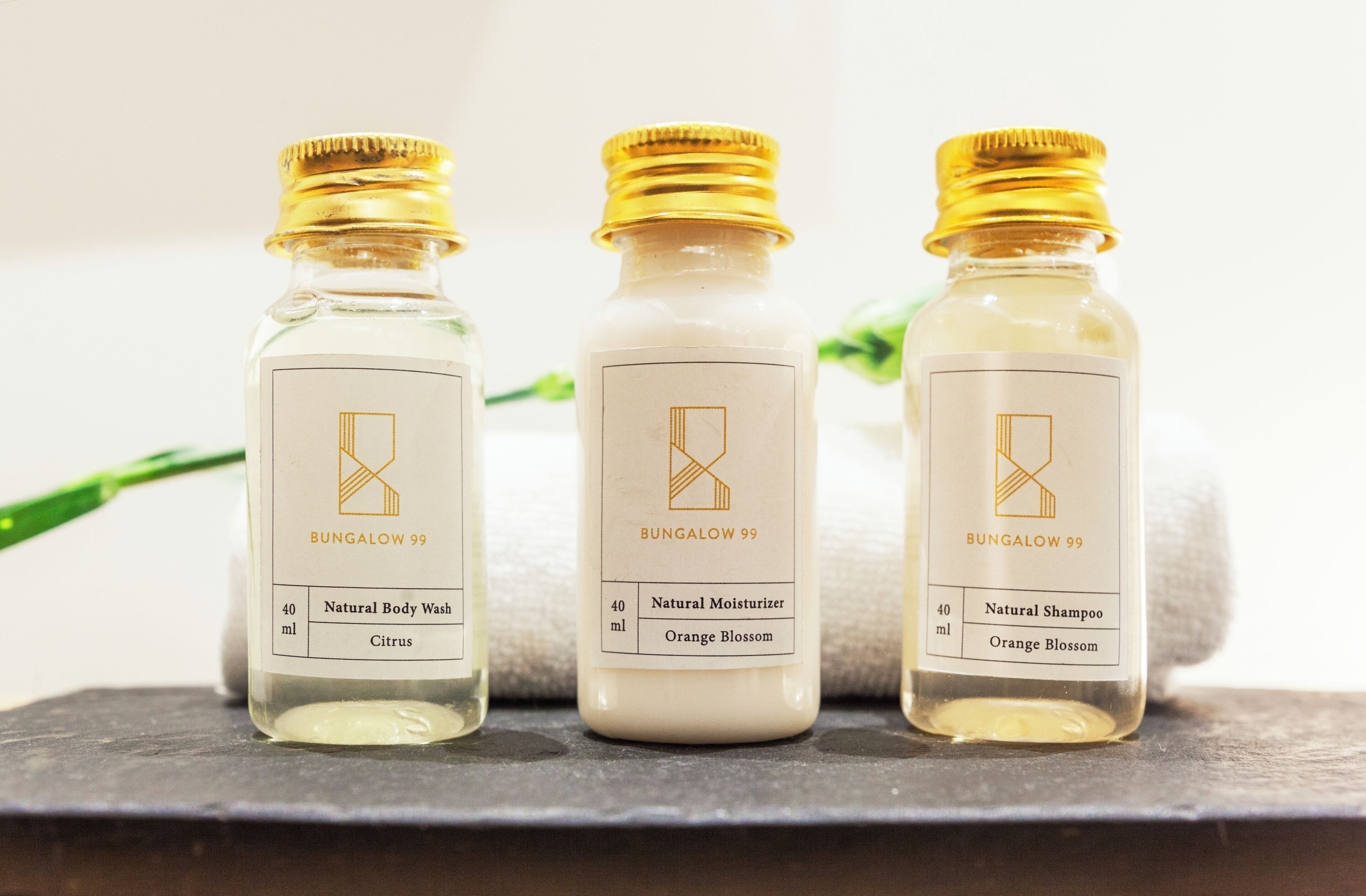 Três embalagens de produtos de cuidados corporais Bungalow 99 sobre uma superfície de pedra, com uma toalha e folhas verdes ao fundo.