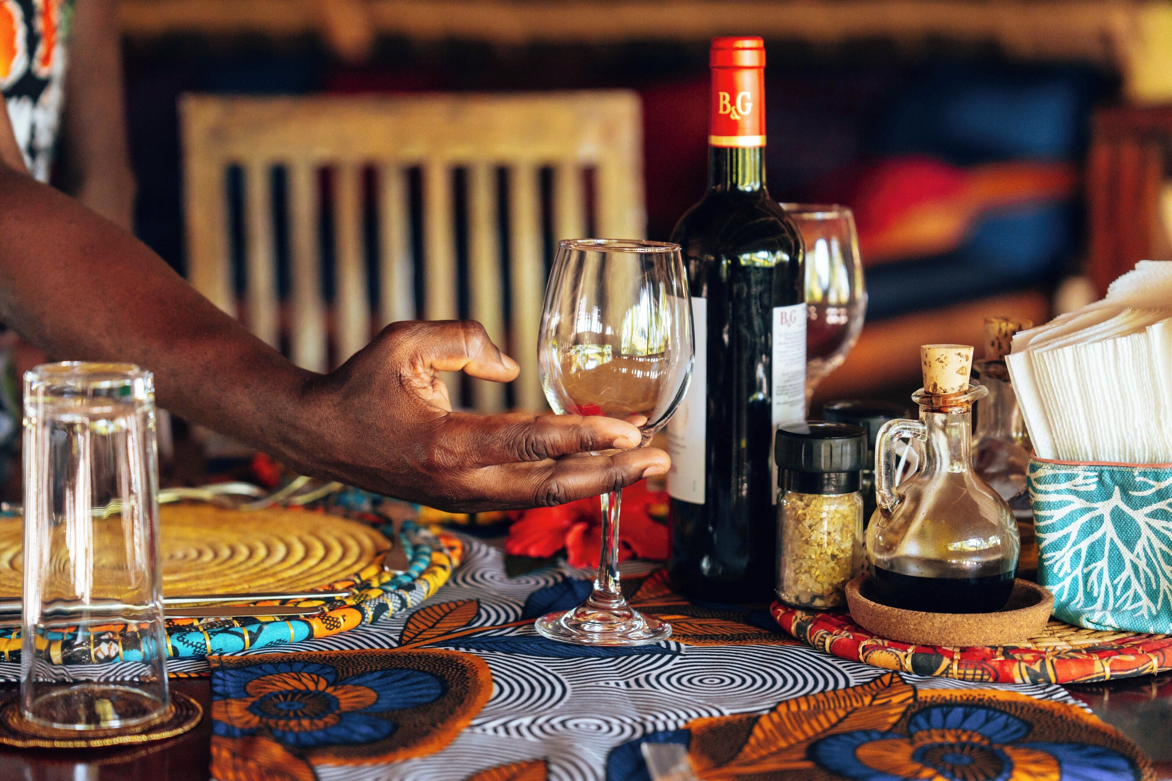 Uma pessoa segurando uma taça de vinho, uma garrafa de vinho e um saca-rolhas sobre uma mesa com toalha de mesa estampada.