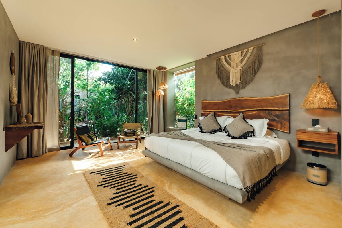 Quarto moderno com uma cama grande, cabeceira de madeira, vista para o verde através de amplas janelas e um espaço de estar acolhedor com uma poltrona e um tapete.