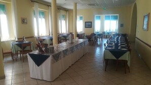 Um amplo salão de jantar com uma longa mesa coberta por um tecido branco e um caminho de mesa azul. Várias mesas menores com toalhas verdes estão dispostas ao redor da mesa principal. O ambiente possui paredes amarelas, janelas grandes com cortinas brancas e piso de azulejos.