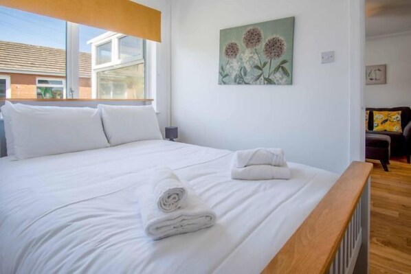 Uma cama bem arrumada com lençóis brancos, um quadro de flores na parede e uma janela com vista para edifícios.