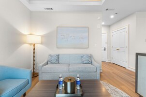 Uma sala de estar moderna com um sofá azul claro, uma poltrona combinando, uma mesa de centro com garrafas de água, um abajur e um quadro com pássaros na parede.