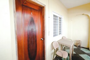 Quarto com porta de madeira marcada com o número 1, janela com grades de metal, mesa com duas cadeiras e uma cadeira de vime pequena.