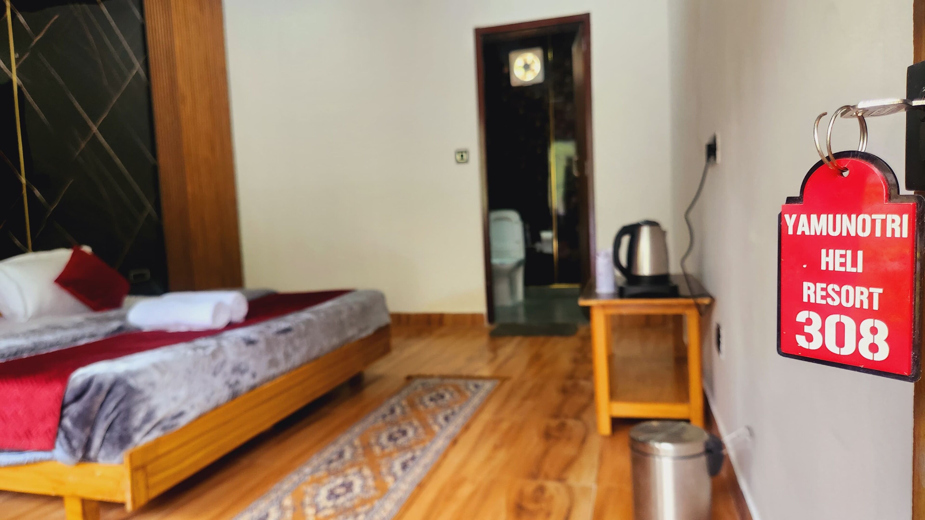 Quarto com cama, mesa de cabeceira, chaleira e placa vermelha indicando 'Yamunotri Heli Resort 308'.