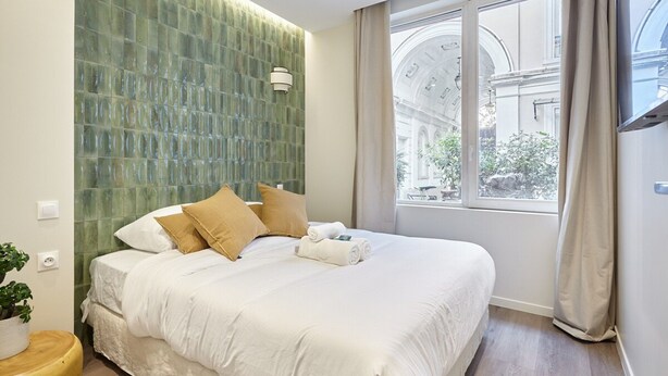 Quarto de hotel com uma cama grande, parede verde com textura, janela com vista para um prédio e um vaso de planta sobre um banquinho amarelo.