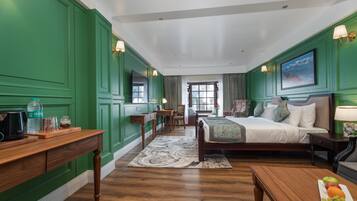 Quarto de hotel com paredes verdes, cama de madeira, escrivaninha com cafeteira e vista para o exterior pela janela.