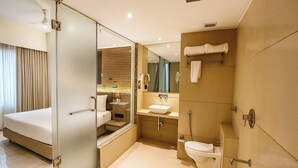 Quarto de hotel moderno com cama, divisória de vidro, banheiro com pia, vaso sanitário e chuveiro.