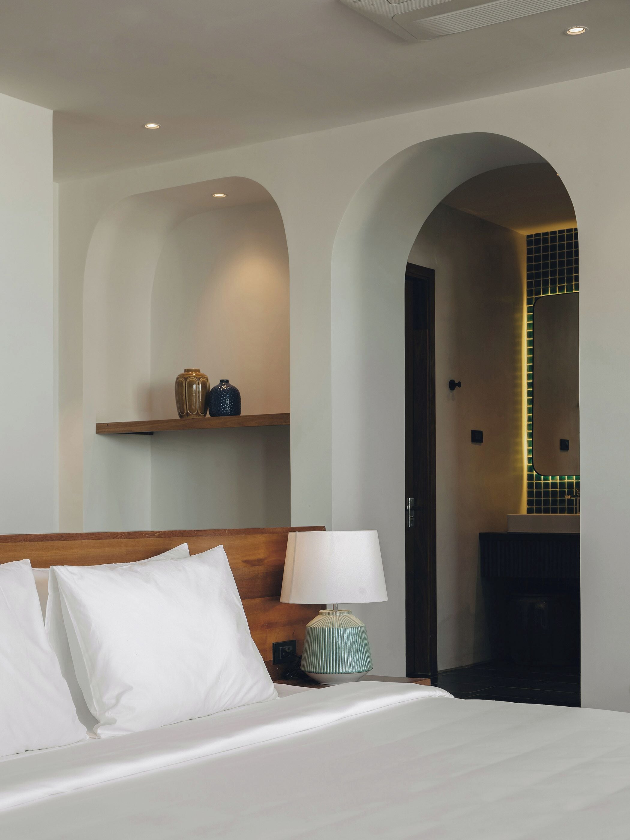 Quarto de hotel com cama, mesa de cabeceira, luminária e prateleira com objetos decorativos.