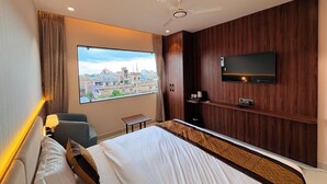 Quarto de hotel com cama, televisão fixada na parede, ventilador de teto, janela com vista para a cidade e uma mesinha de cabeceira com abajur.
