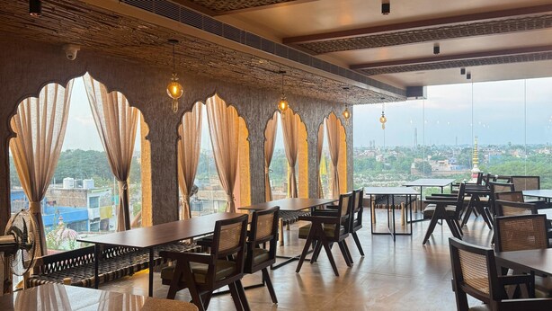 Restaurante com teto alto, design de teto estampado e grandes janelas com cortinas. Mesas e cadeiras dispostas para refeições.