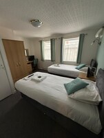 Quarto de hotel com duas camas de solteiro, uma escrivaninha com cadeira, guarda-roupa e janela com cortinas.