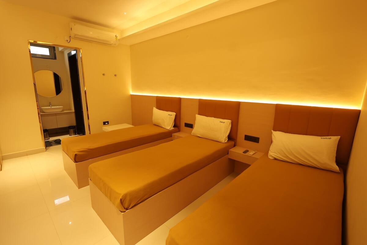 Quarto de hotel com três camas de solteiro, uma mesa de cabeceira com luminária e banheiro com pia e espelho.