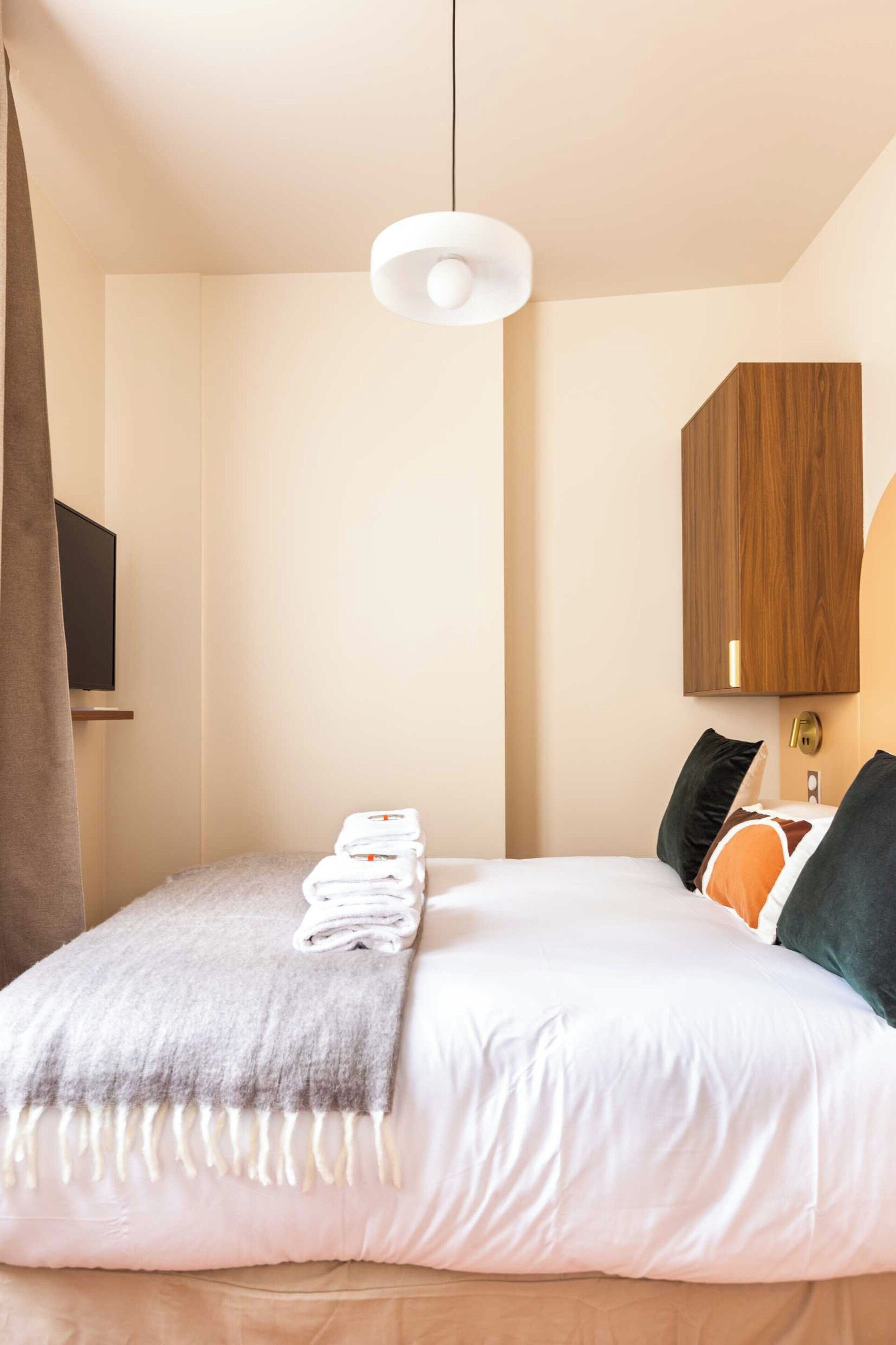 Cama bem arrumada com edredom branco, manta cinza e toalhas dobradas. Existe um armário de madeira acima da cama e uma televisão montada na parede à esquerda.