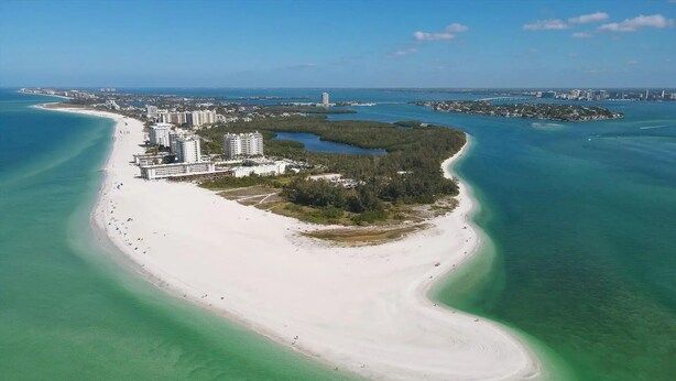 Vista aérea de uma área costeira com uma praia de areia branca e curvilínea, um conjunto denso de edifícios e um mar azul cristalino.