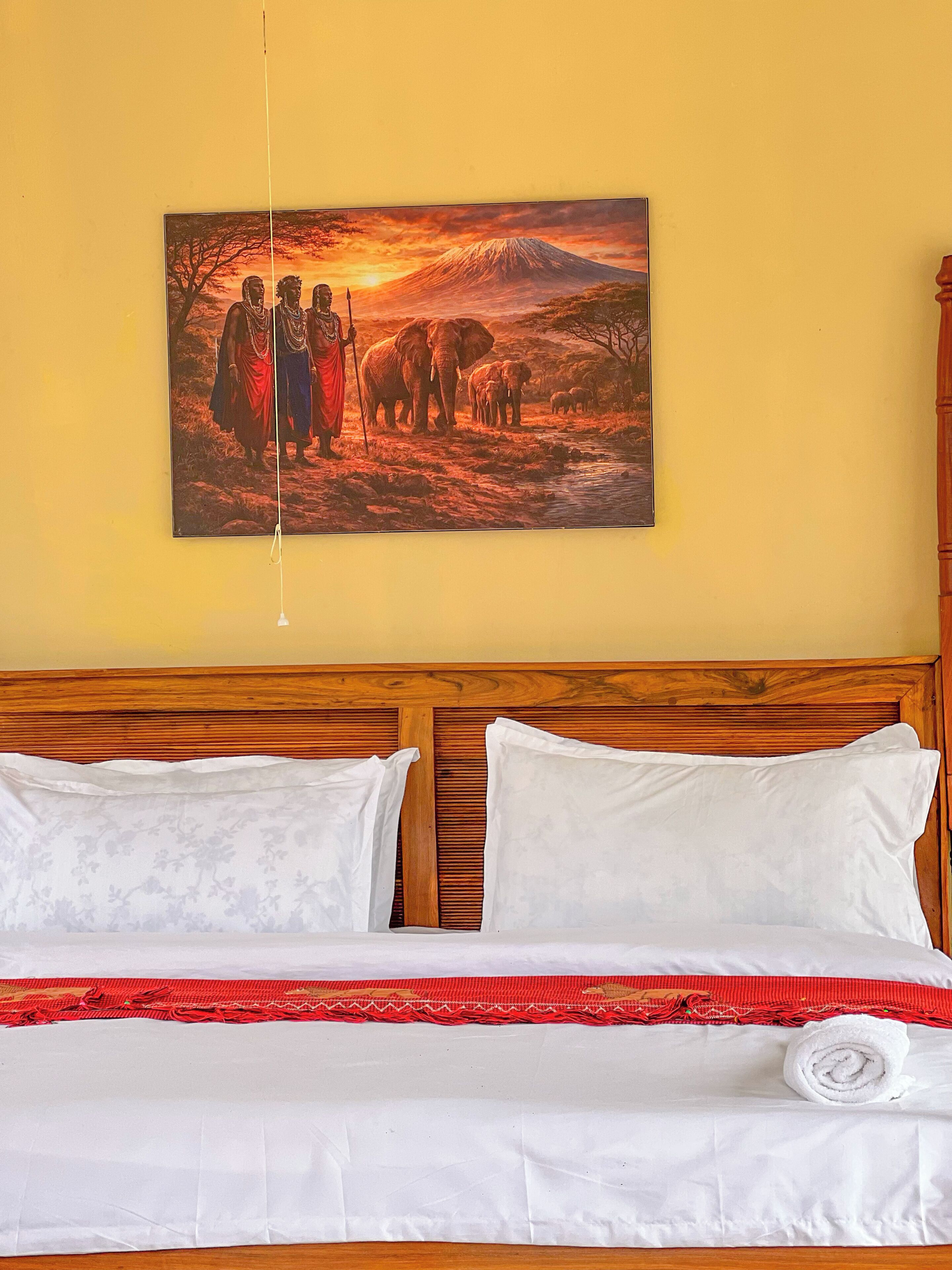 Uma cama com lençóis brancos e uma borda decorativa vermelha, uma cabeceira de madeira e um quadro na parede retratando um grupo de pessoas e elefantes.