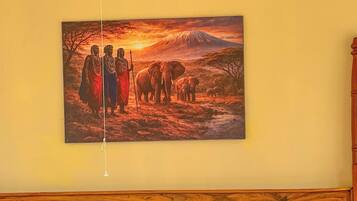 Uma cama com lençóis brancos e uma borda decorativa vermelha, uma cabeceira de madeira e um quadro na parede retratando um grupo de pessoas e elefantes.