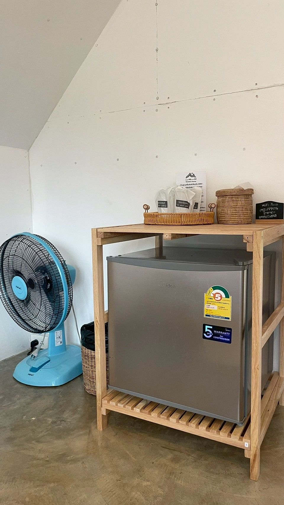 Uma cozinha compacta com geladeira de aço inoxidável, um ventilador azul e uma mesa de madeira com cesta e placa.