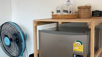 Uma cozinha compacta com geladeira de aço inoxidável, um ventilador azul e uma mesa de madeira com cesta e placa.