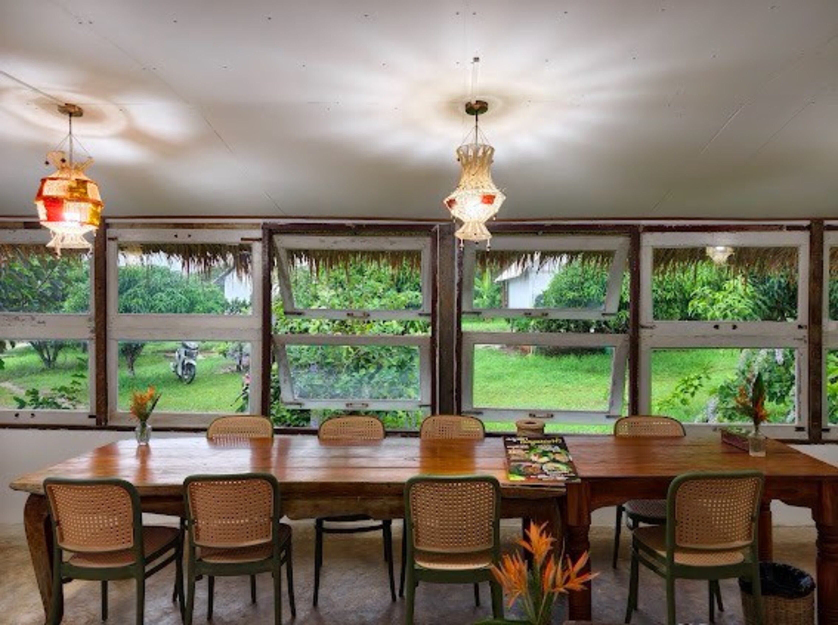 Uma sala de jantar com uma mesa de madeira comprida, cadeiras de vime e grandes janelas com vista para um jardim.