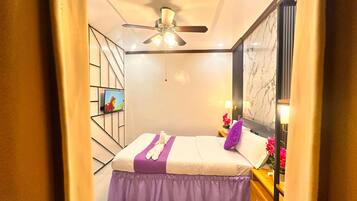 Quarto de hotel com cama, ventoinha no teto, televisão fixada na parede e mesa de cabeceira com abajur.