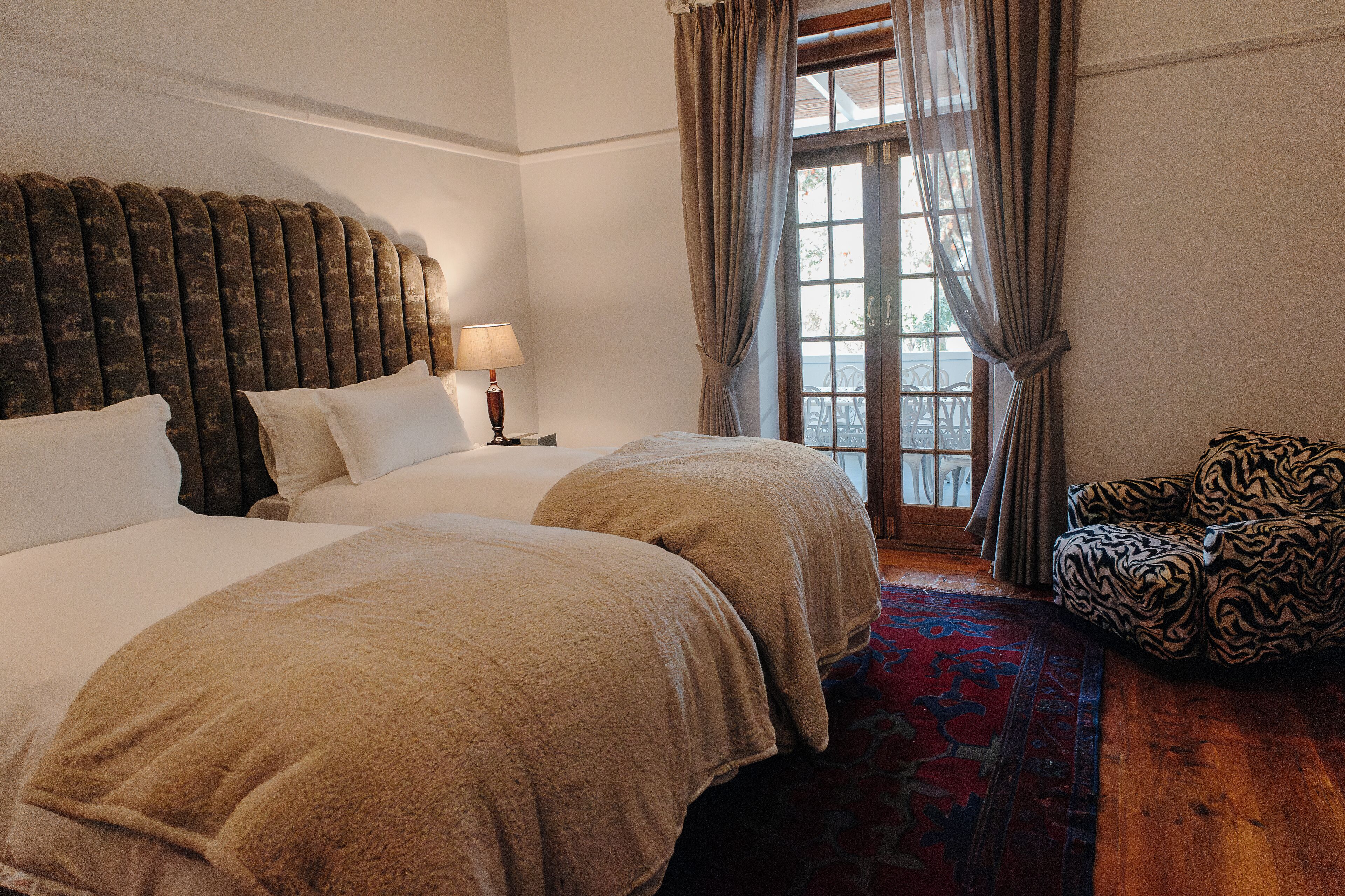 Quarto de hotel com duas camas, uma poltrona estampada, uma janela com cortinas e uma mesa de cabeceira com abajur.