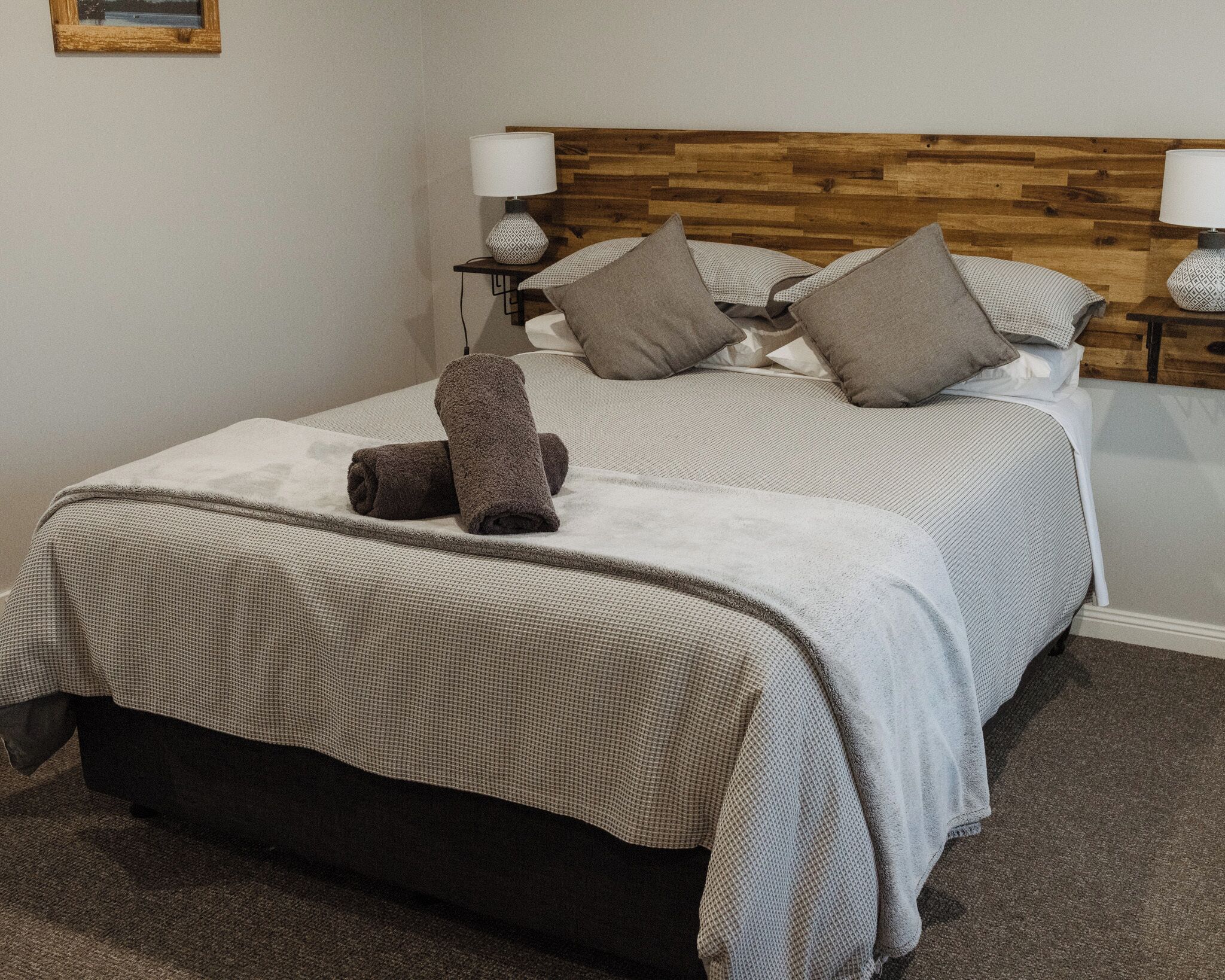 Cama bem arrumada com colcha xadrez, dois travesseiros e uma toalha enrolada. Há duas mesinhas de cabeceira com abajures e uma cabeceira de madeira.